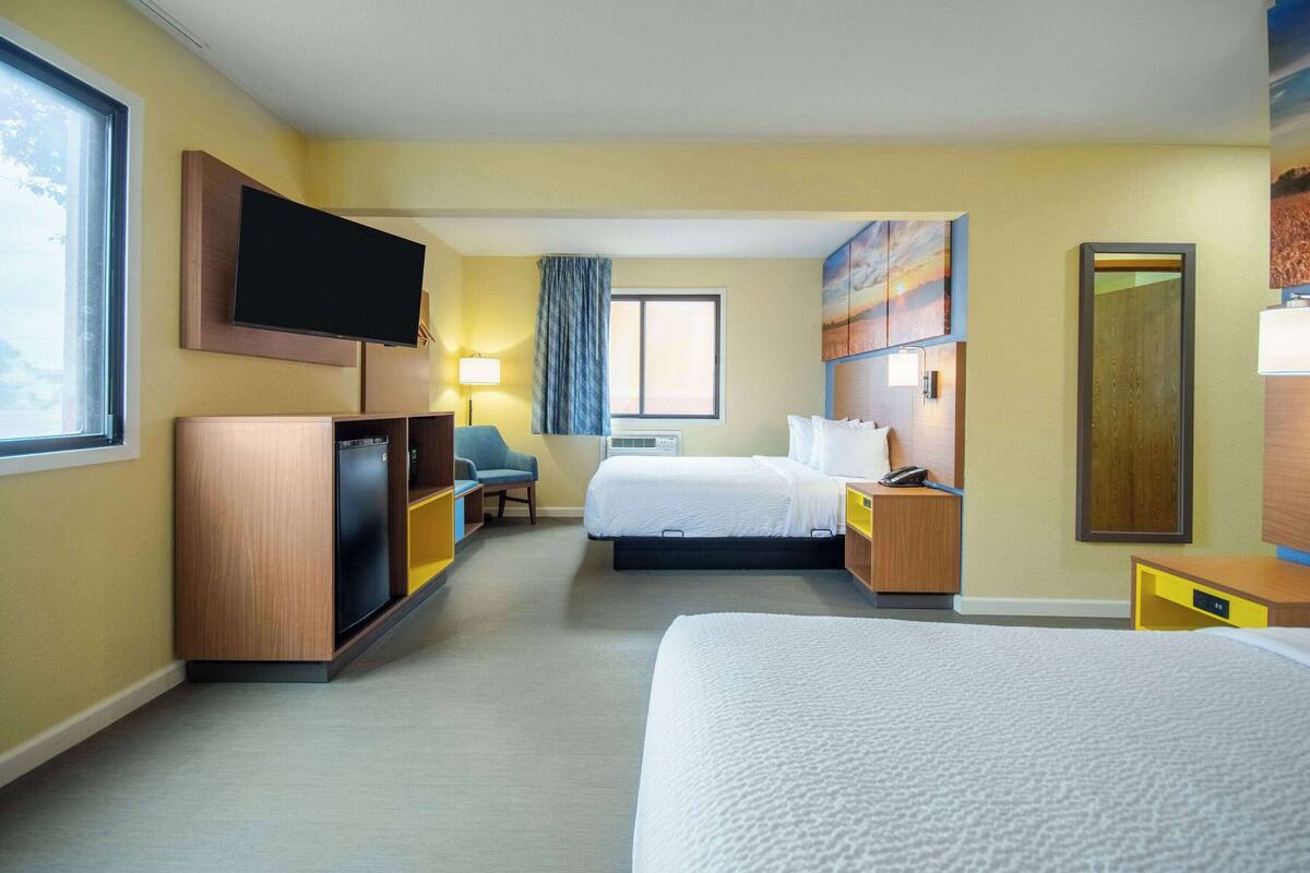 Quarto de hotel com uma cama grande, uma cama menor, uma televisão fixada na parede, uma mesa com cadeira e uma janela com cortinas.
