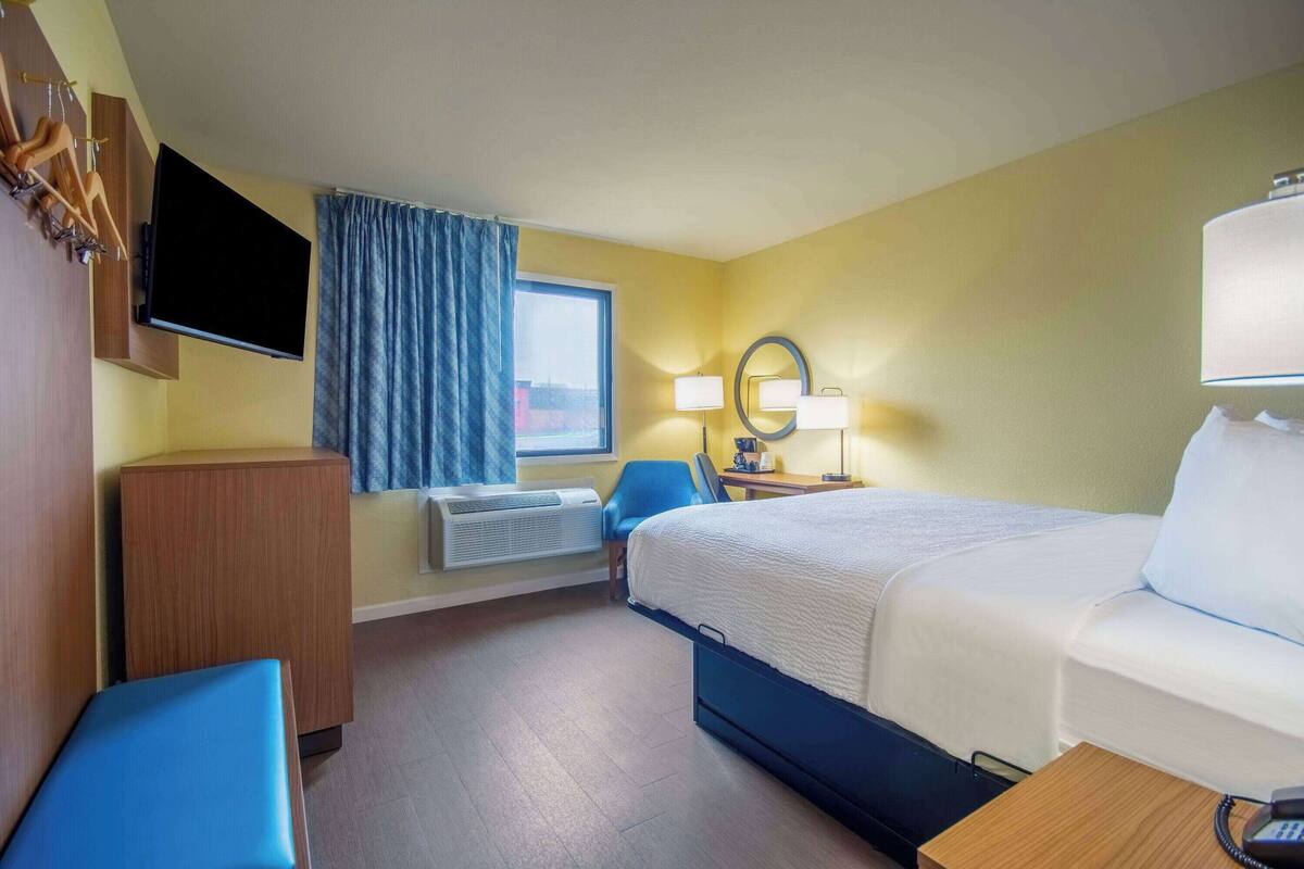 Quarto de hotel com cama, escrivaninha, cadeira, televisão, janela com cortinas, luminária e mesa de cabeceira.