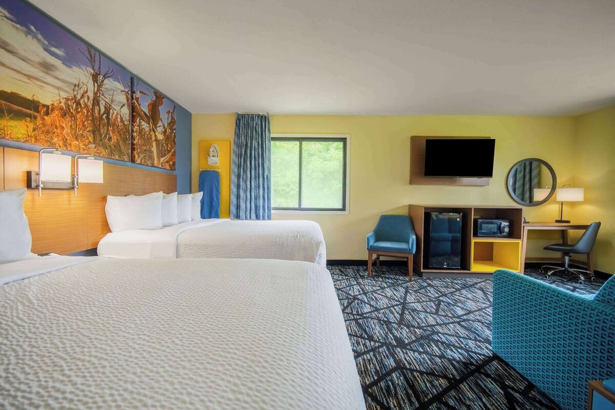 Quarto de hotel com duas camas, uma escrivaninha, uma cadeira, uma televisão e uma janela grande.