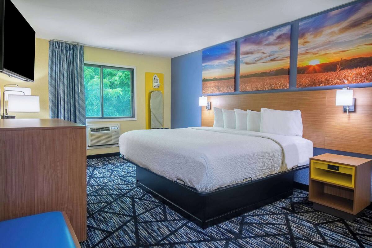 Quarto de hotel com cama, escrivaninha, cadeira, televisão, janela com cortinas e parede com pinturas de pôr do sol.