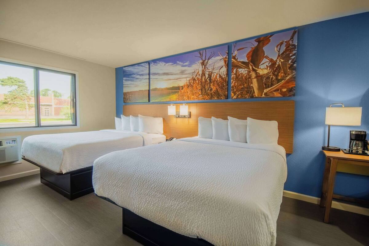 Quarto de hotel com duas camas, uma janela grande, um quadro na parede e uma mesa de cabeceira com abajur e cafeteira.