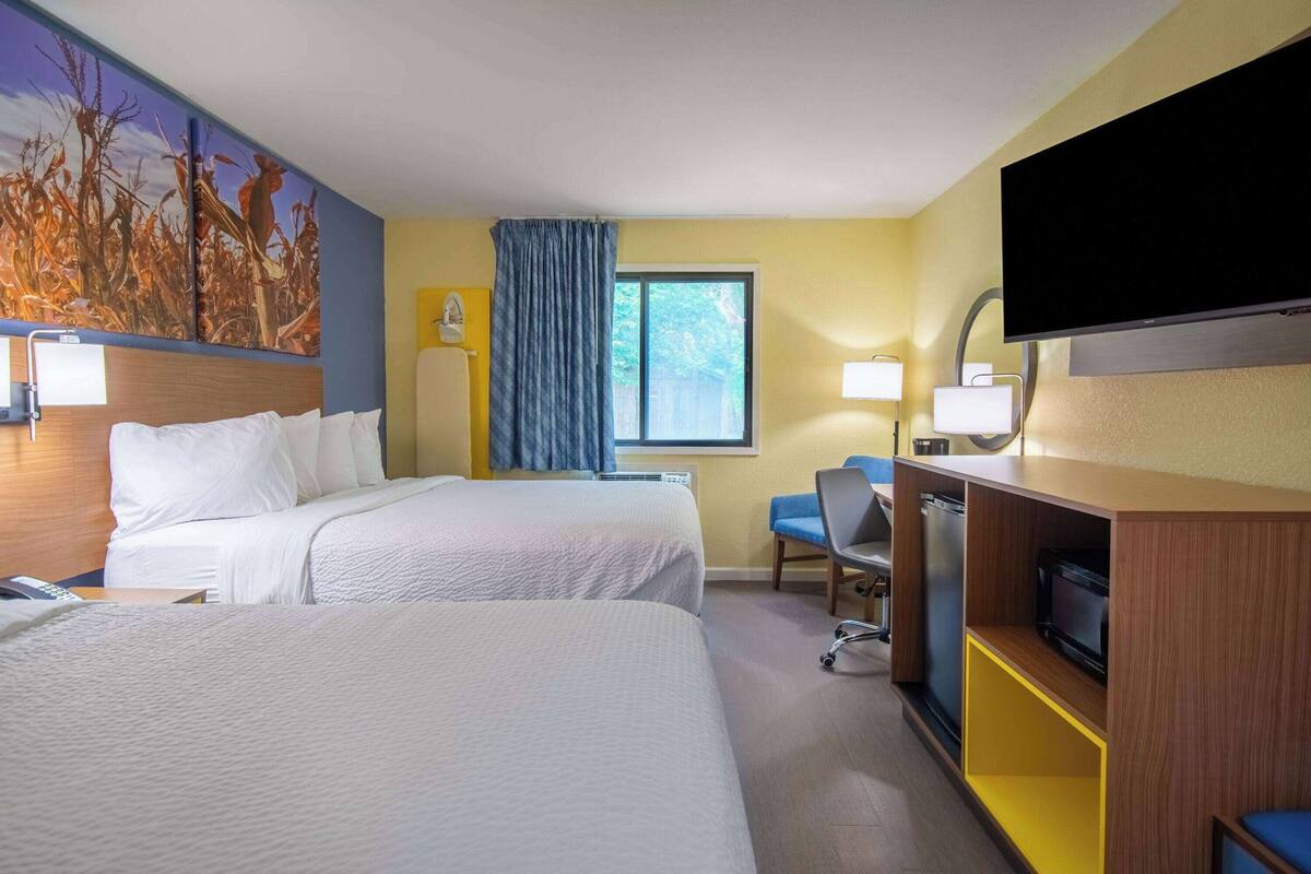 Quarto de hotel com duas camas, uma escrivaninha, uma cadeira, uma televisão e uma janela com cortinas.