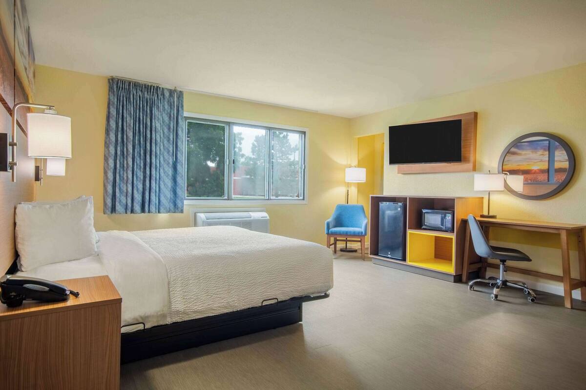 Quarto de hotel com cama, mesa de cabeceira com telefone, escrivaninha com cadeira, televisão, micro-ondas e uma grande janela com cortinas.