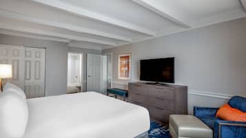 Quarto de hotel com uma cama grande, televisão, cômoda e área de estar com uma cadeira azul e um travesseiro laranja.