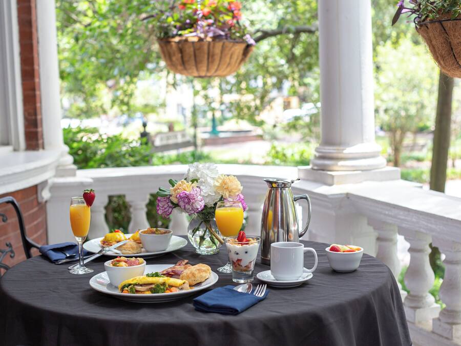 Mesa de café da manhã posta com diversos pratos, um jarro e um vaso de flores em uma varanda com vista para o verde.