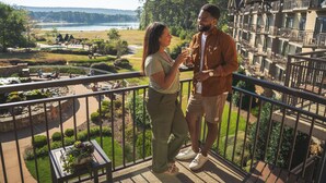 Um casal numa varanda com vista para um jardim ajardinado, um corpo de água e edifícios ao fundo.