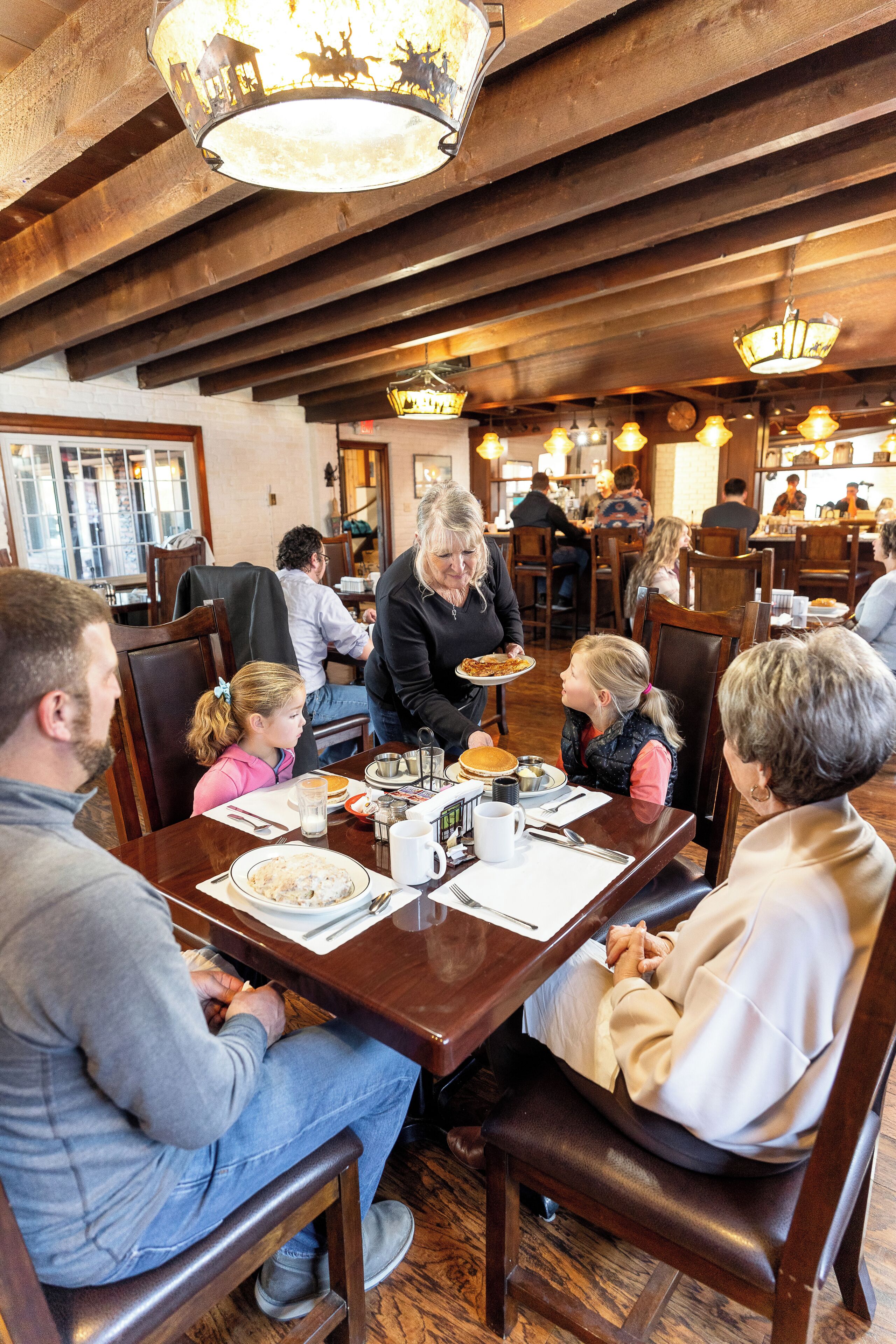 Interior de restaurante com mesas e cadeiras de madeira, um lustre e uma parede de tijolos.