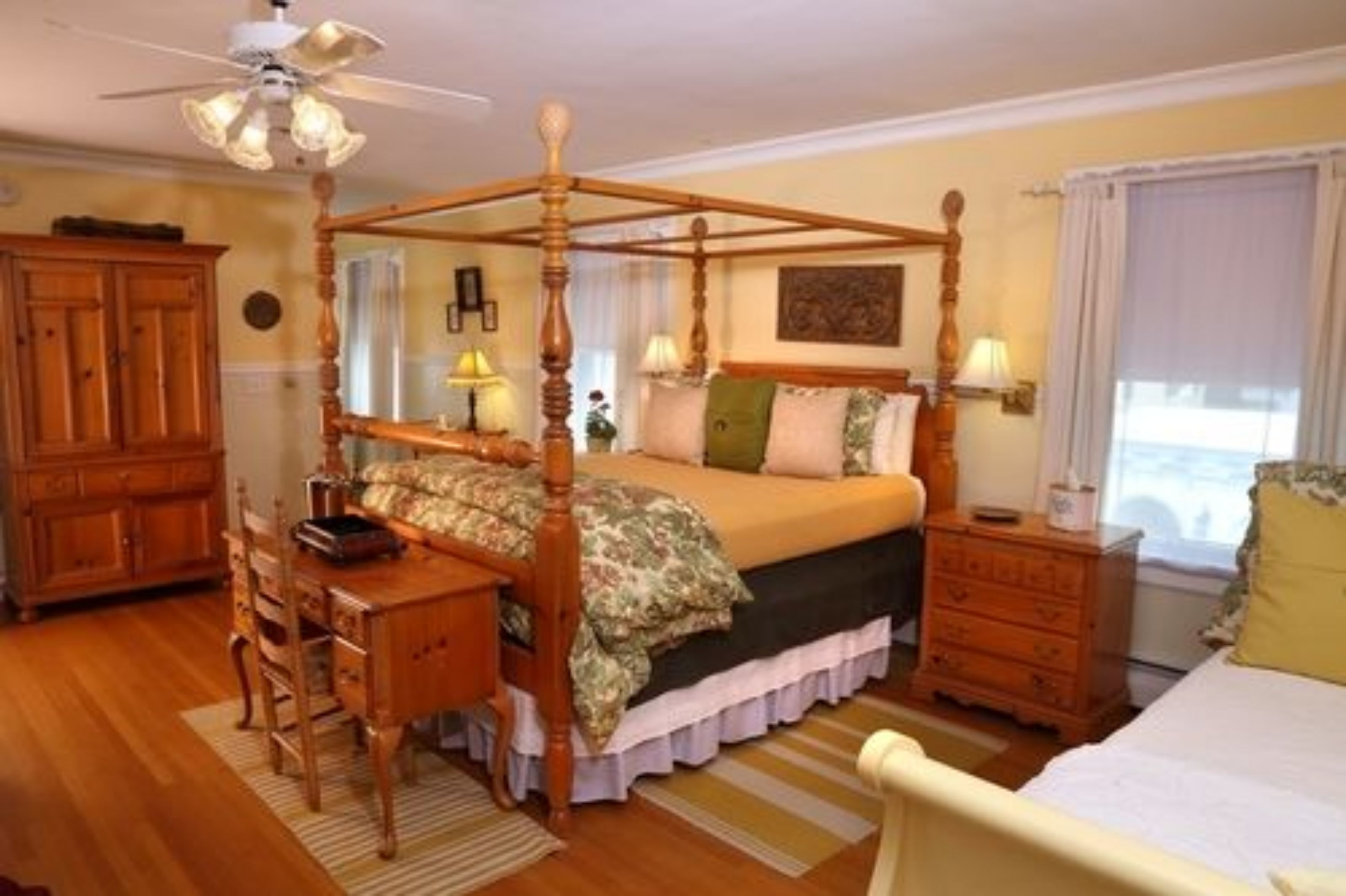 Uma cama de dossel com um céu de madeira, uma escrivaninha com cadeira, um aparador e uma janela com cortinas.