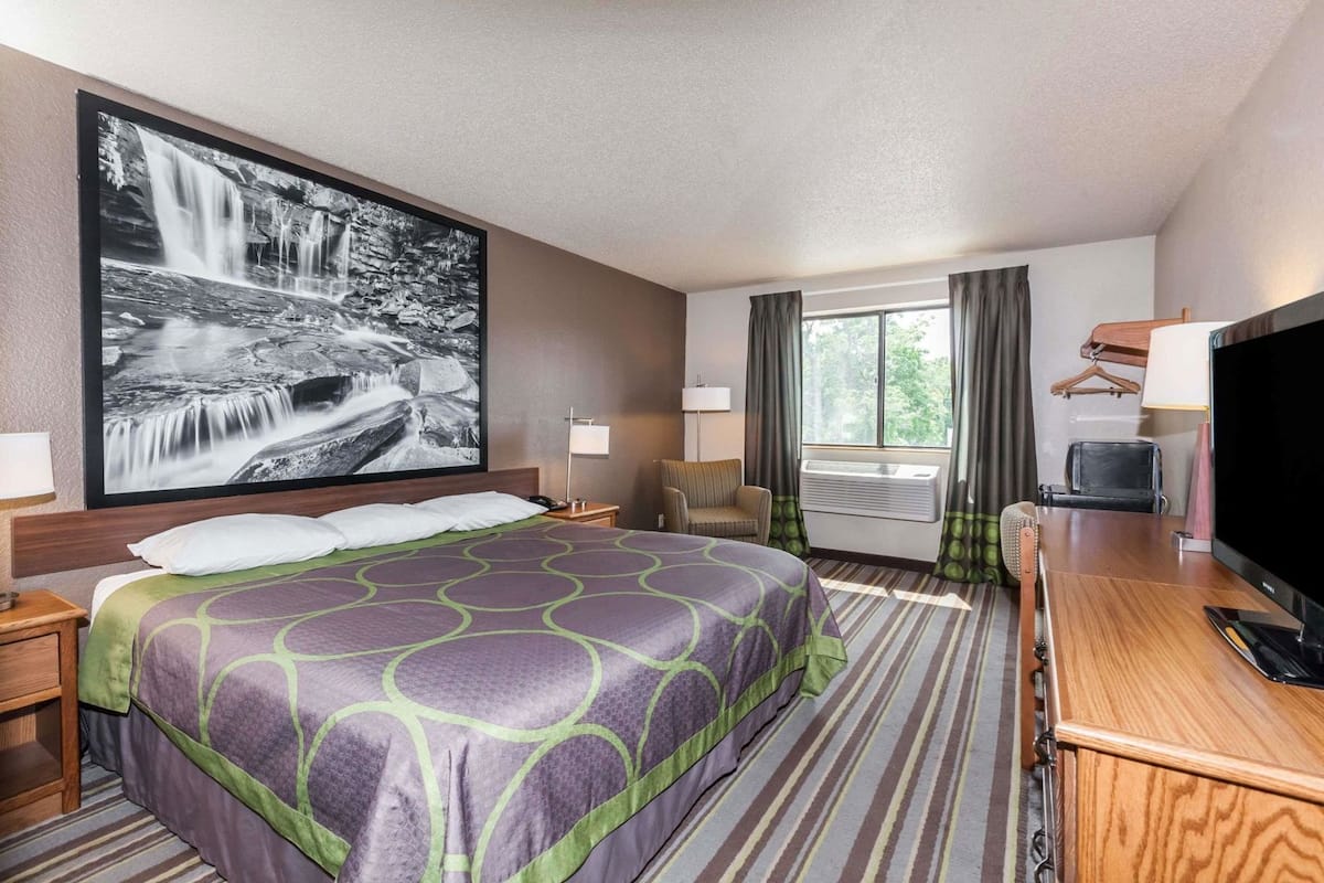 Quarto de hotel com uma cama grande, uma escrivaninha, televisão, janela com cortinas e um quadro na parede de uma cascata.