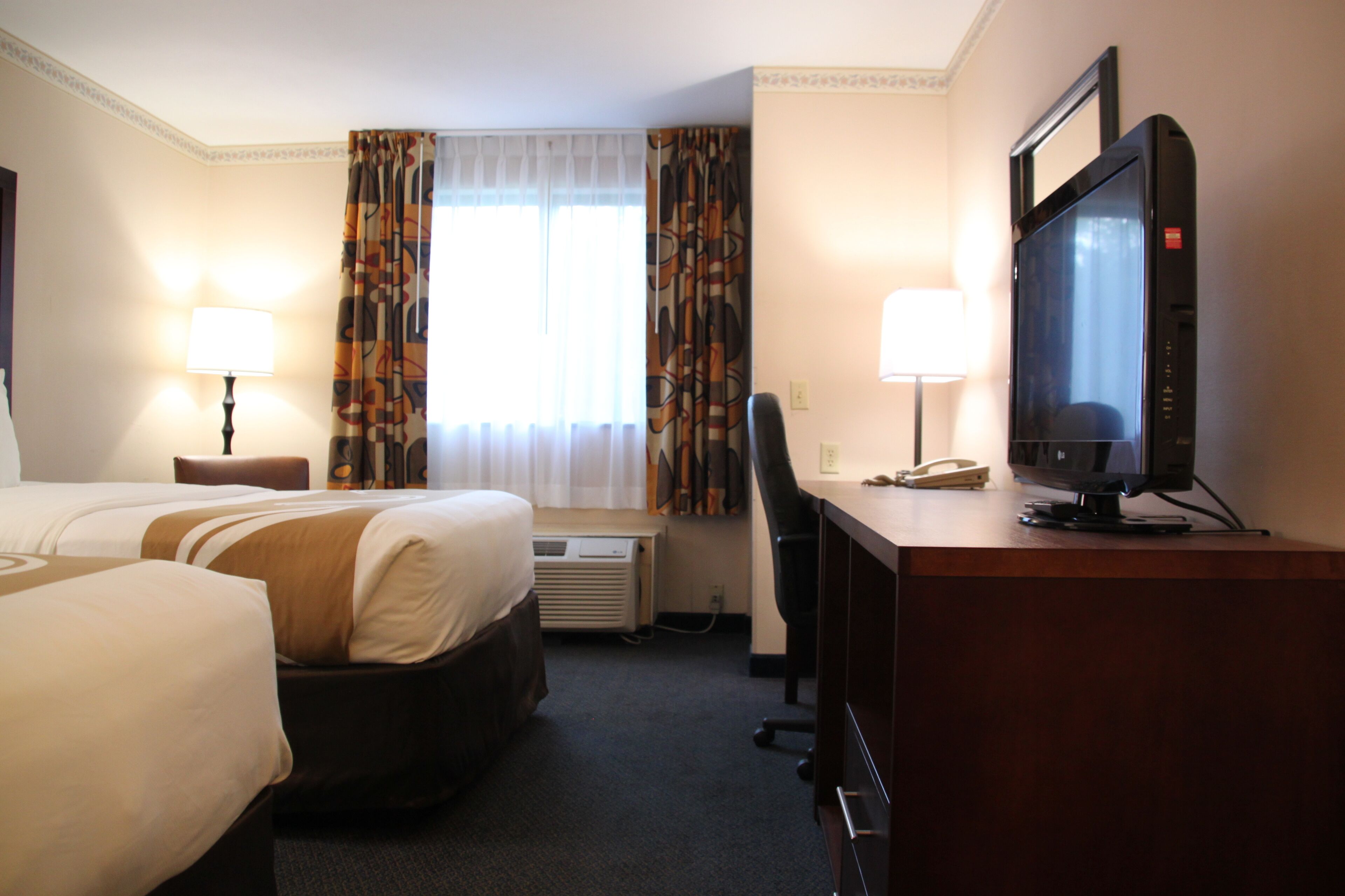 Quarto de hotel com duas camas, uma escrivaninha com computador, uma cadeira e uma janela com cortinas.