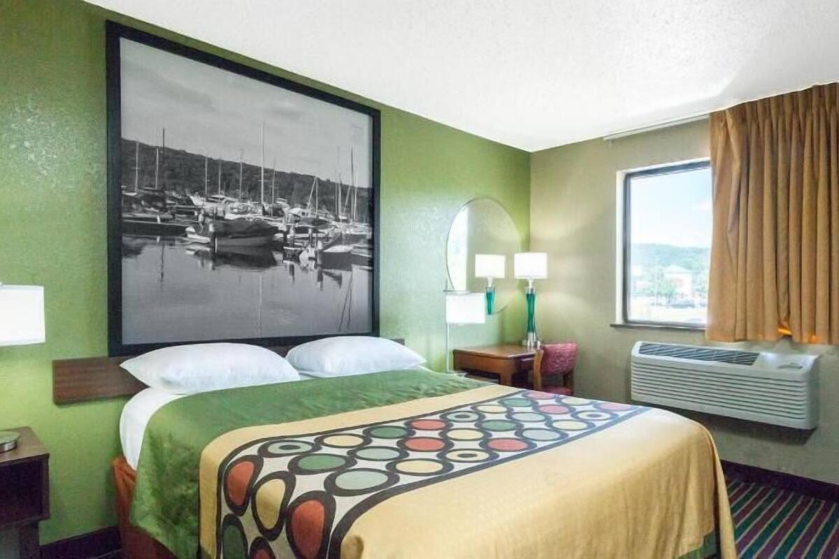 Quarto de hotel com uma cama, duas luminárias de cabeceira, um quadro grande emoldurado, uma janela com cortinas e um aparelho de ar condicionado.