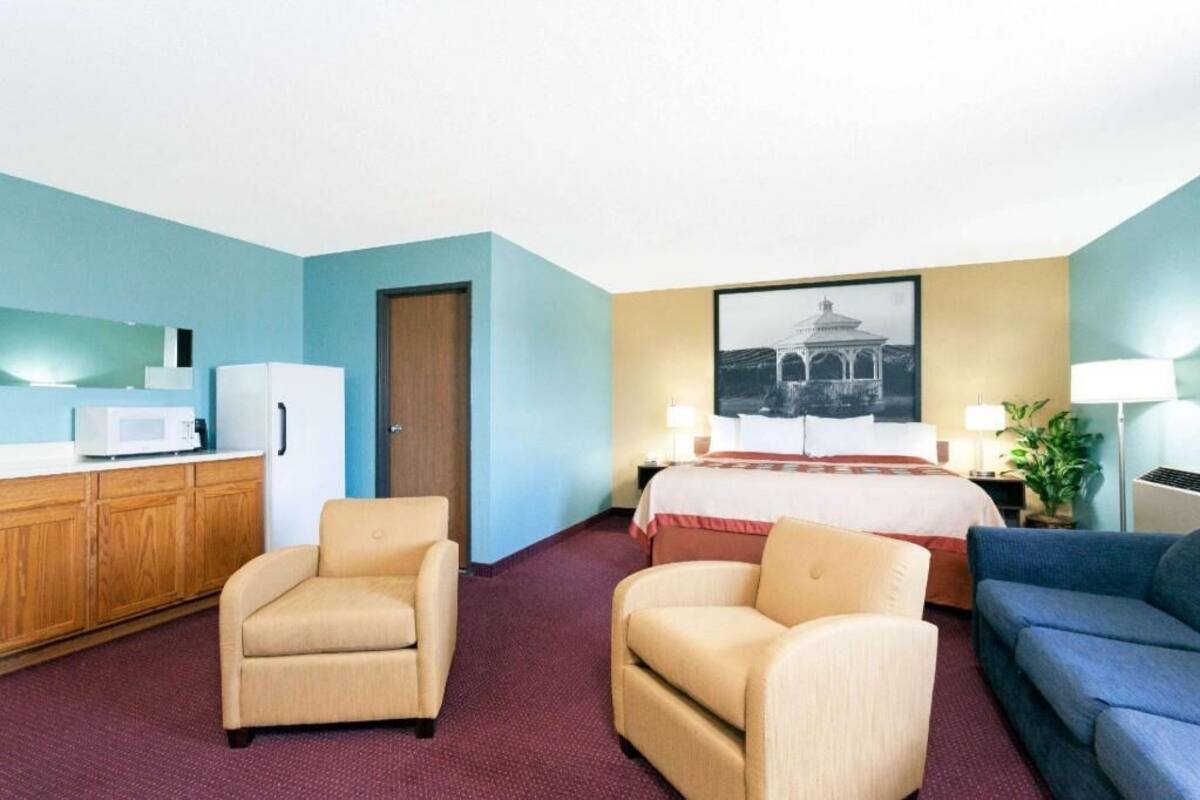 Quarto de hotel com cama, duas poltronas, um sofá, micro-ondas, frigorífico, mesa de cabeceira com abajur e um quadro na parede.