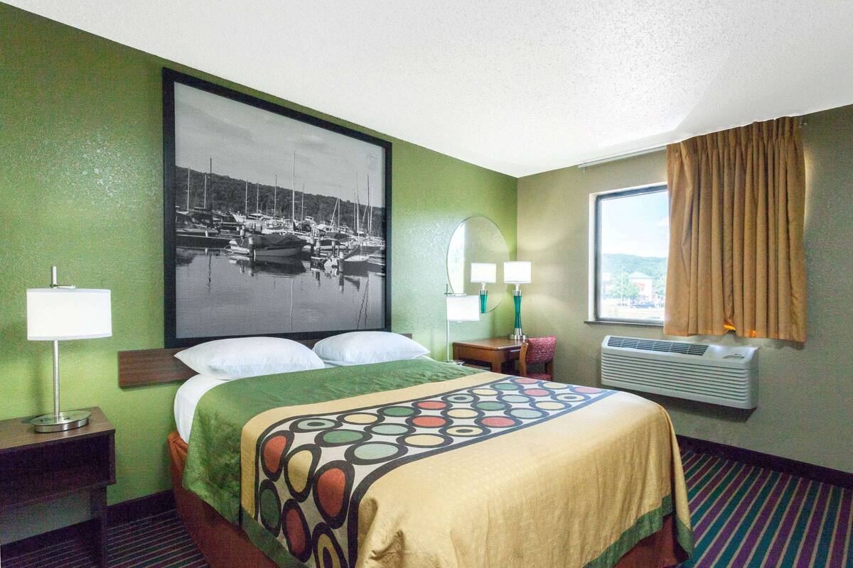 Quarto de hotel com cama, mesa de cabeceira, escrivaninha, cadeira, janela com cortinas, ar condicionado e espelho fixado na parede.