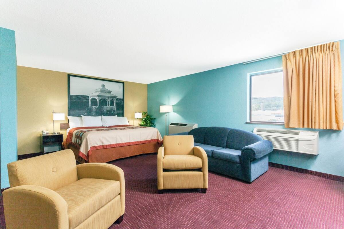 Quarto de hotel com cama, duas poltronas, um sofá, uma mesinha de cabeceira, uma luminária, uma janela com cortinas e um ar condicionado fixado na parede.