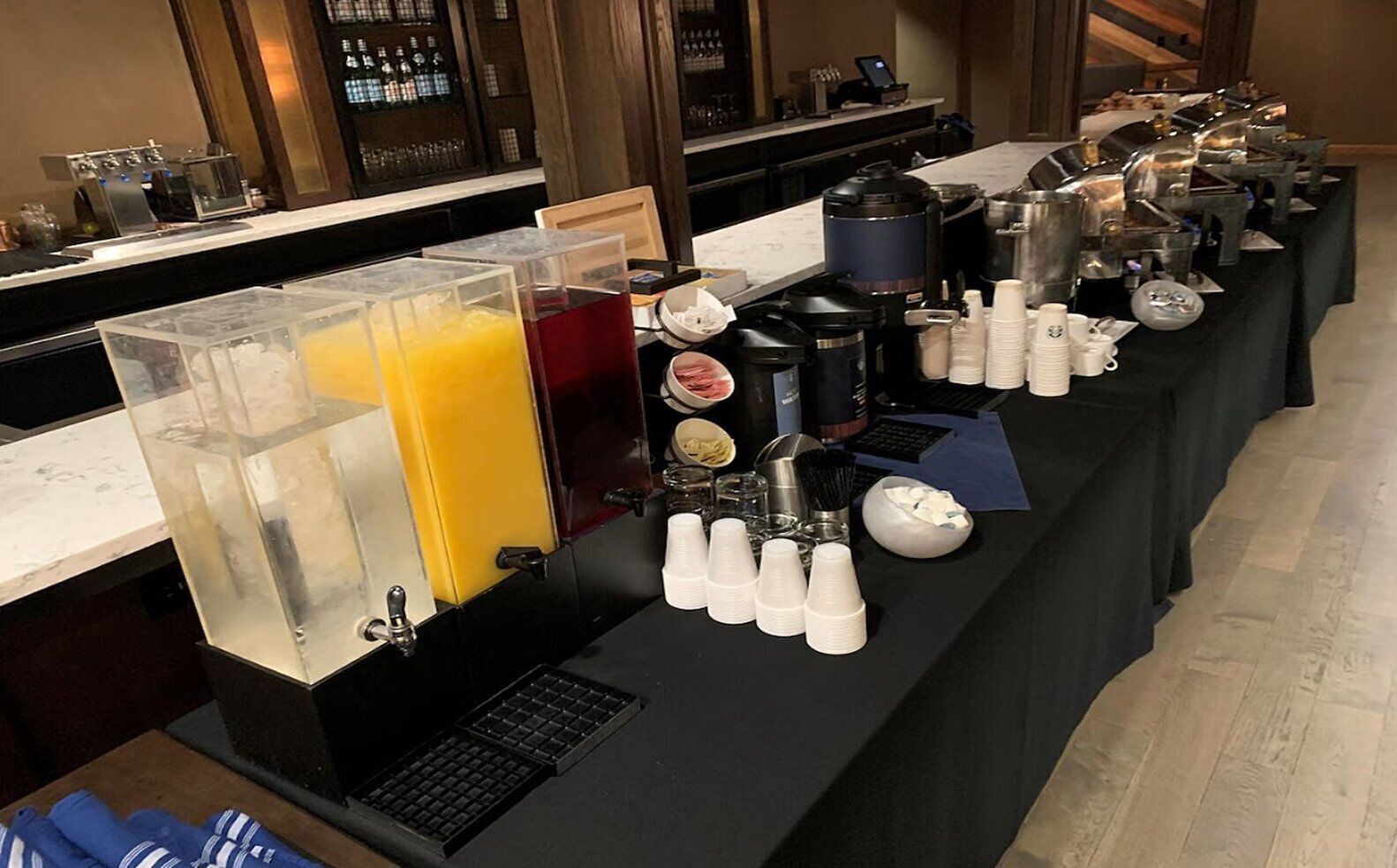 Uma mesa de pequeno-almoço com dispensador de sumo, máquinas de café e diversos copos e recipientes.