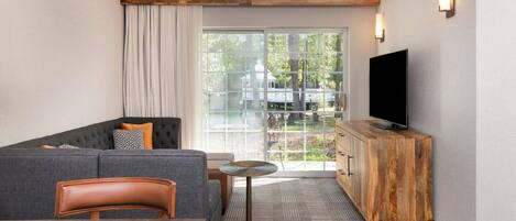 Quarto de hotel moderno com um sofá cinzento, um armário de madeira, uma TV e uma varanda com porta de vidro.