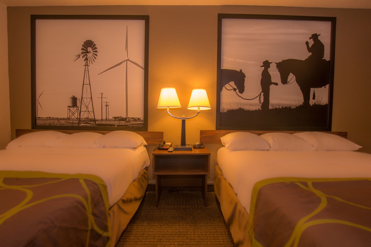 Quarto de hotel com duas camas, uma mesa de cabeceira com abajur e duas fotografias emolduradas na parede.