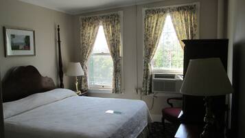 Quarto de hotel com uma cama, duas janelas com cortinas, uma mesa de trabalho com abajur e uma cadeira.