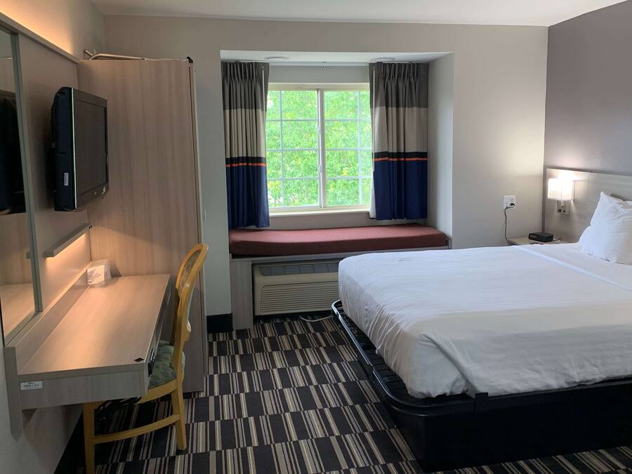 Quarto de hotel com cama, escrivaninha com cadeira, televisão, janela com cortinas e mesa de cabeceira com luminária.