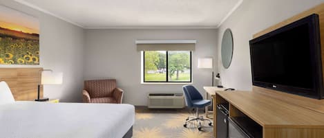 Quarto de hotel com cama, escrivaninha com televisão, cadeira, janela com vista para o verde e espelho fixado na parede.