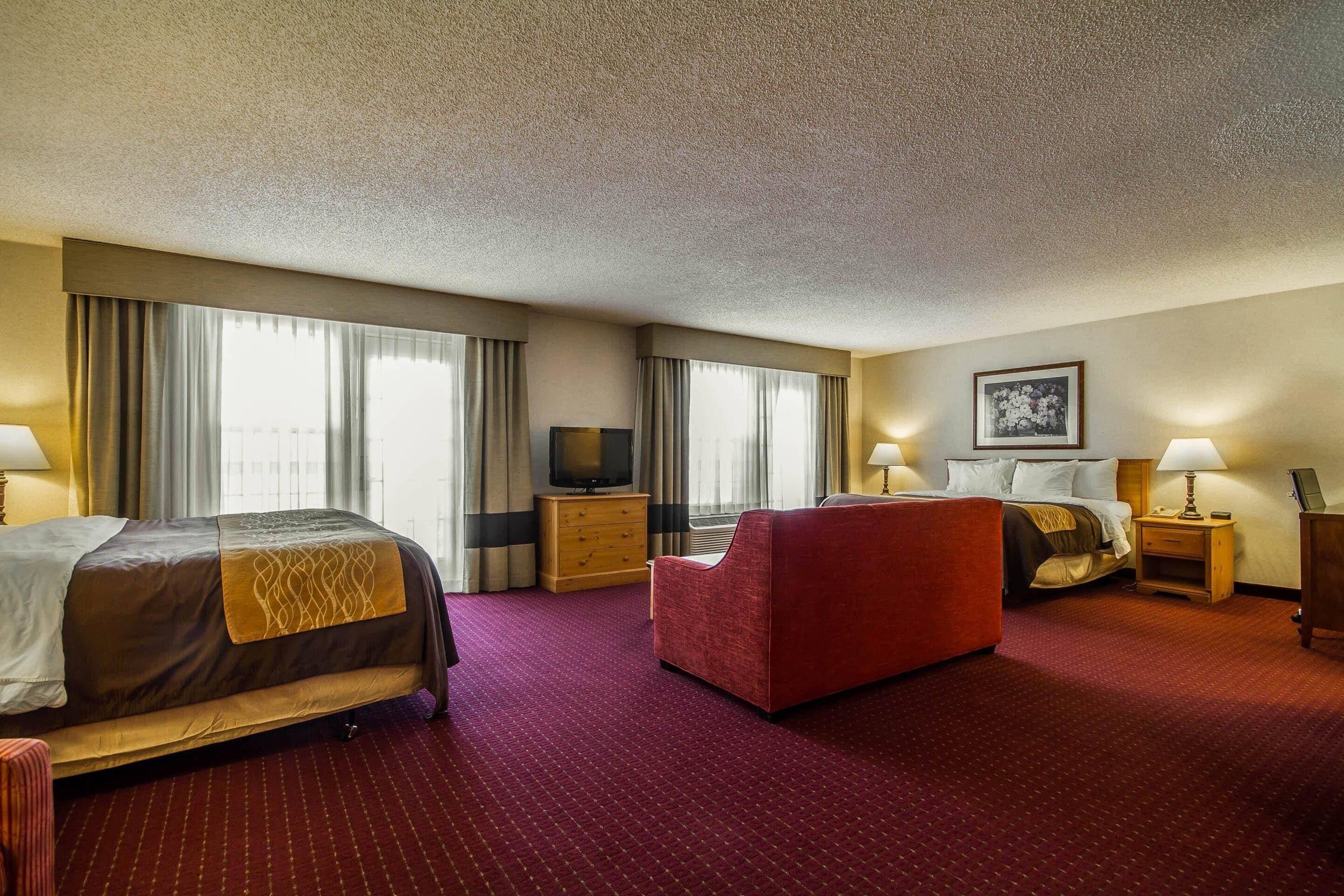 Quarto de hotel com cama, uma poltrona vermelha, televisão, escrivaninha e luminária.