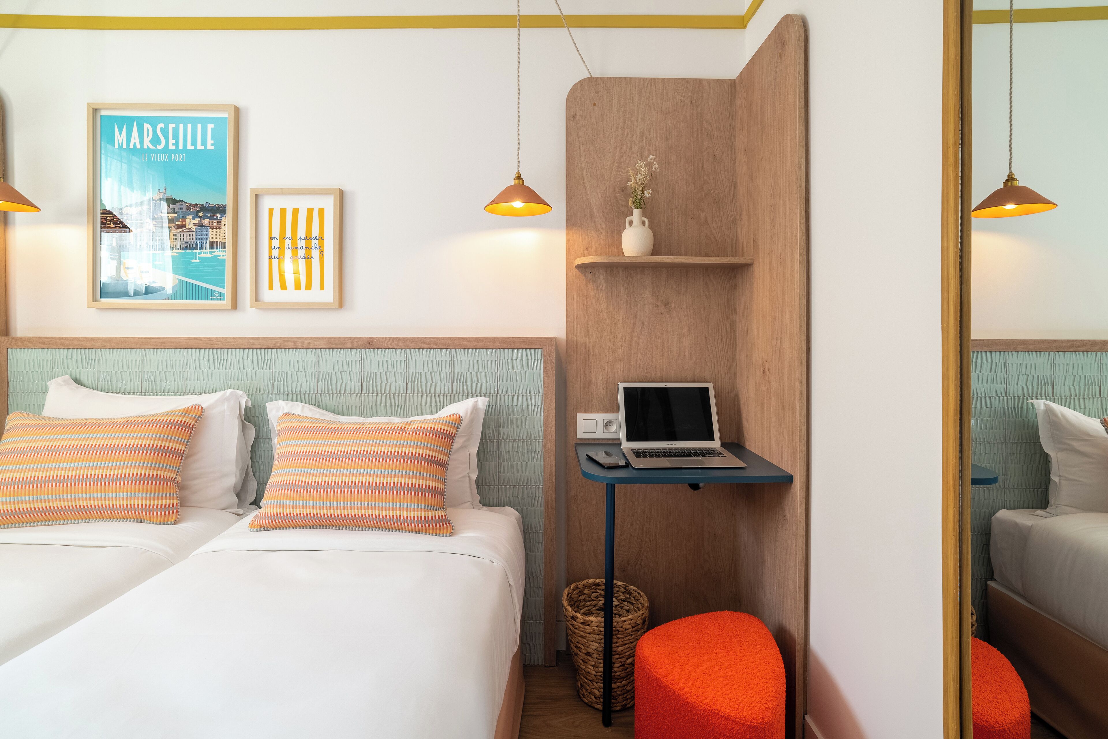 Quarto de hotel com duas camas, um laptop sobre uma mesinha, um quadro emoldurado de Marselha e um espelho.