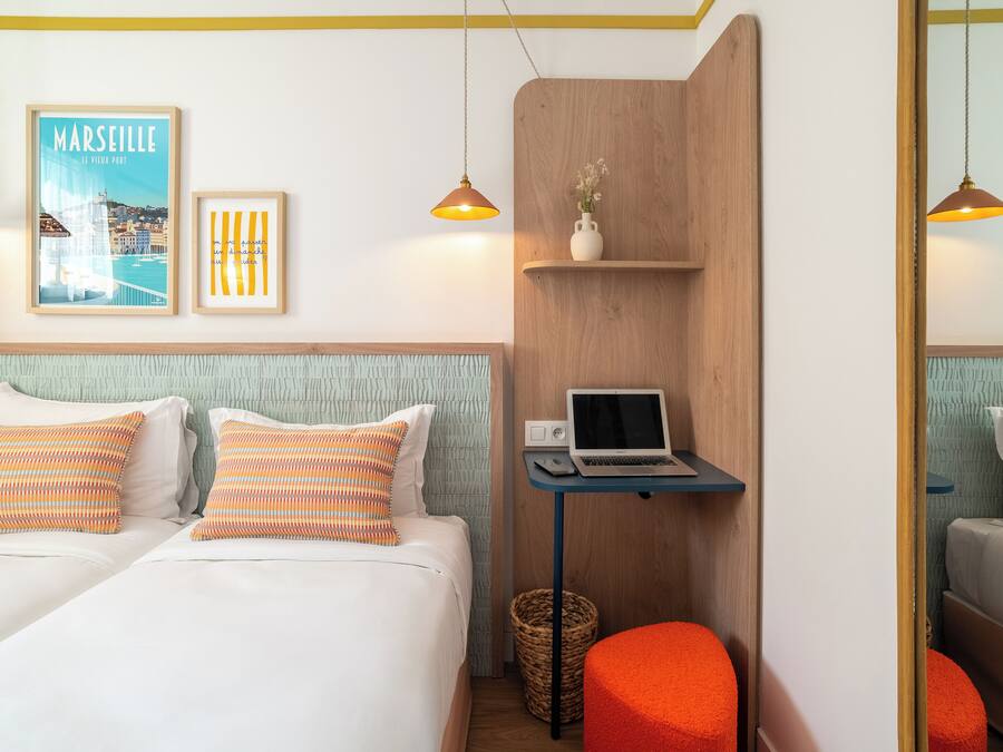 Quarto de hotel com duas camas, um laptop sobre uma mesinha, um quadro emoldurado de Marselha e um espelho.
