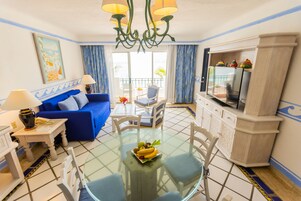 Quarto de hotel com sofá azul, mesa de jantar de vidro com frutas, televisão e varanda com cortinas azuis.