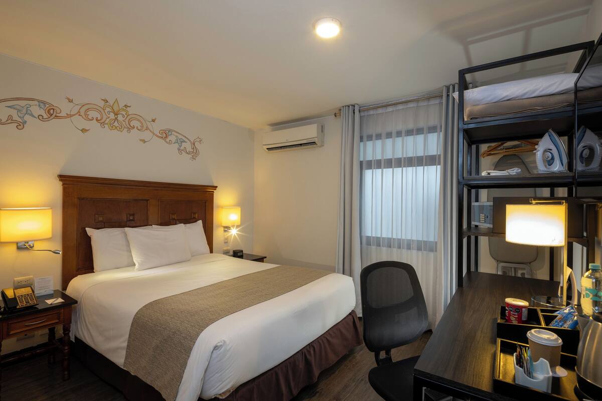 Quarto de hotel com uma cama grande, duas luminárias de cabeceira, uma escrivaninha com cadeira, um espelho e uma prateleira com artigos de higiene pessoal.