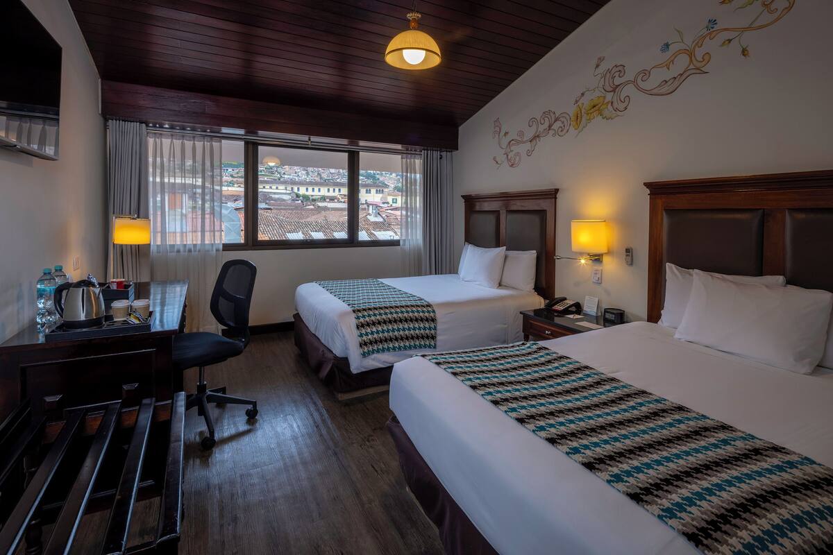 Quarto de hotel com duas camas, uma escrivaninha com cadeira, uma luminária e vista para a cidade pela janela.