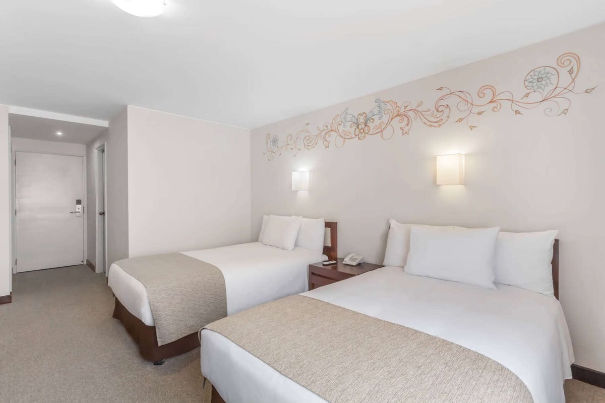 Quarto de hotel com duas camas, uma mesa de cabeceira, uma luminária na parede e uma porta que dá para outro cômodo.