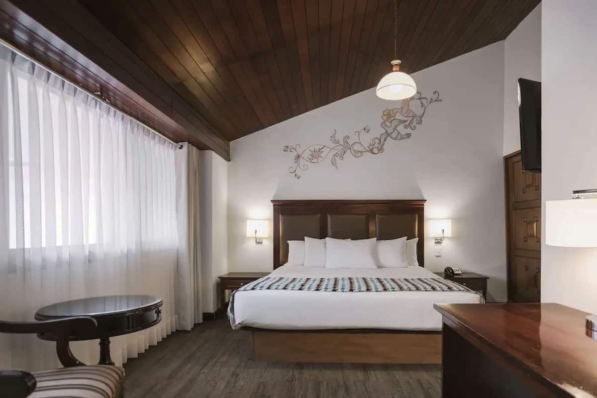 Quarto de hotel com teto de madeira, uma cama grande com roupa de cama branca, uma mesa redonda pequena e uma cadeira.