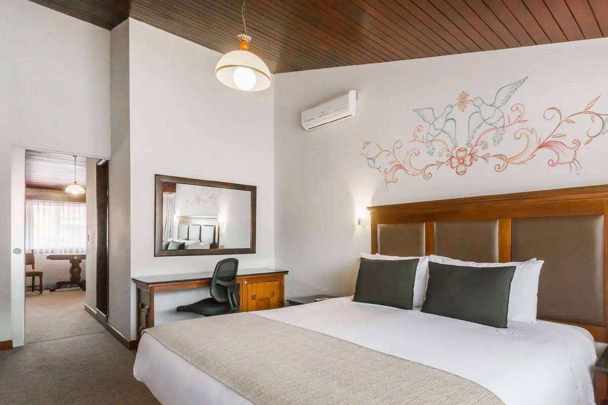 Quarto de hotel com uma cama grande, uma escrivaninha com cadeira, um espelho e um mural decorativo na parede.