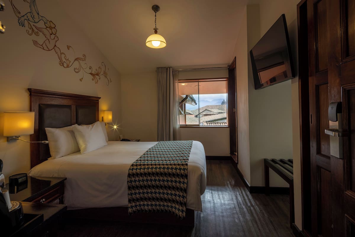 Quarto de hotel com uma cama grande, abajures, cabeceira de madeira, janela com vista e televisão na parede.