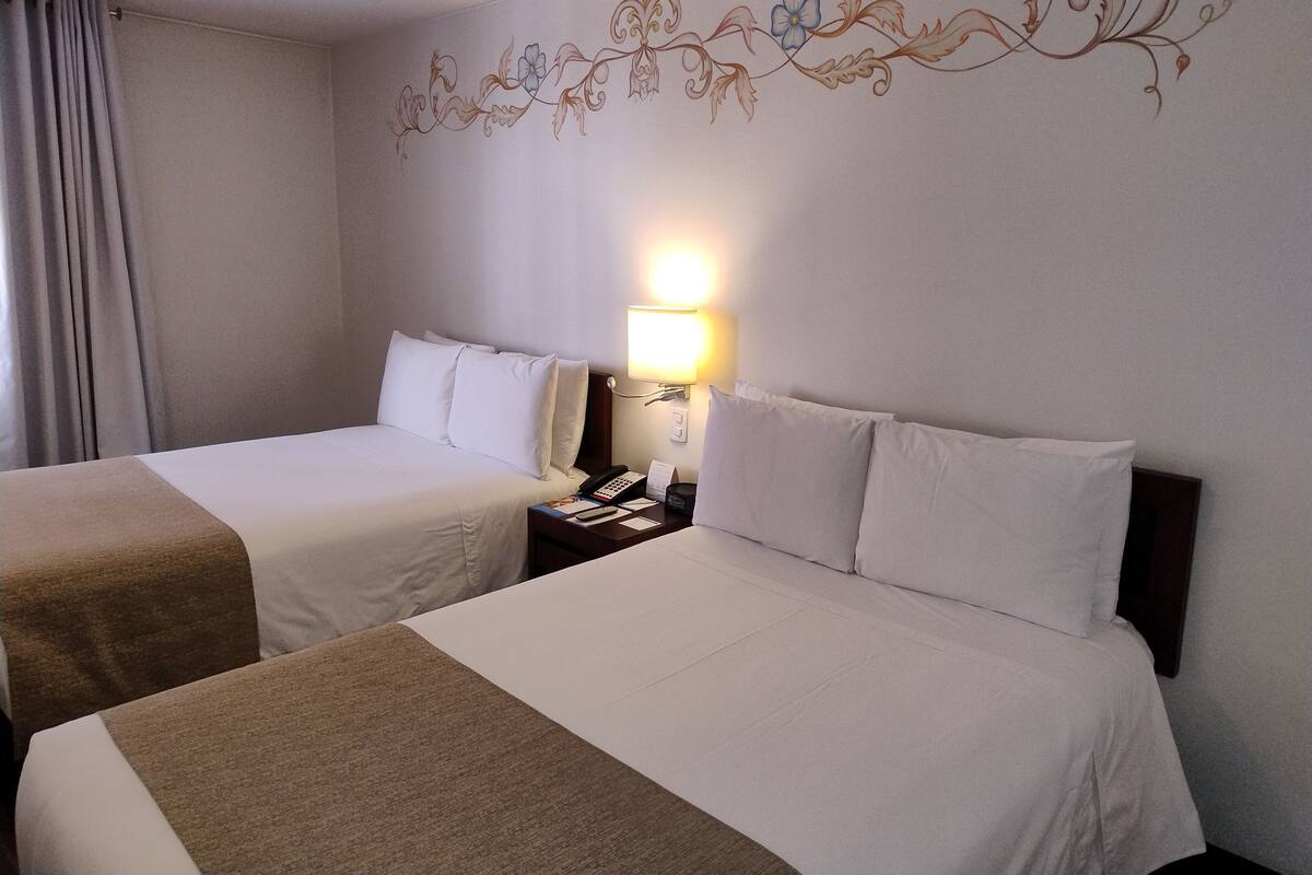 Quarto de hotel com duas camas, uma mesa de cabeceira com abajur e um desenho floral decorativo na parede.