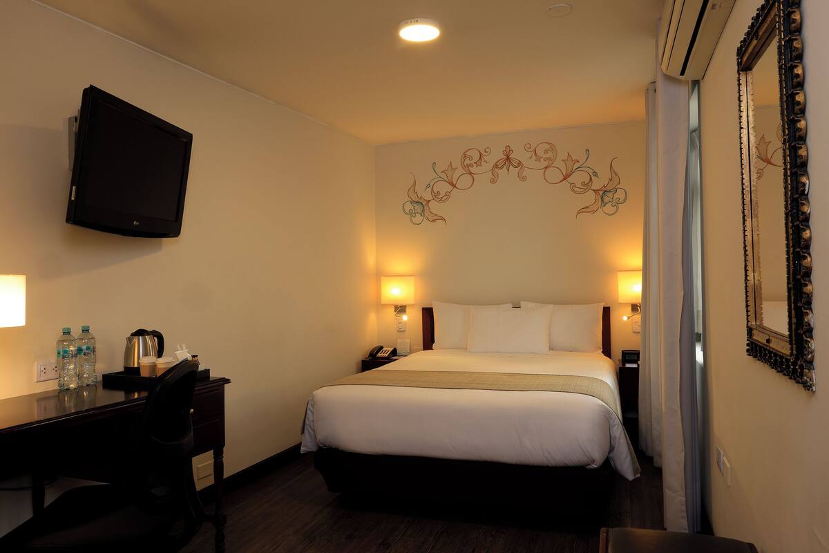 Quarto de hotel com cama, mesa de cabeceira, escrivaninha, cadeira, televisão e decoração na parede.