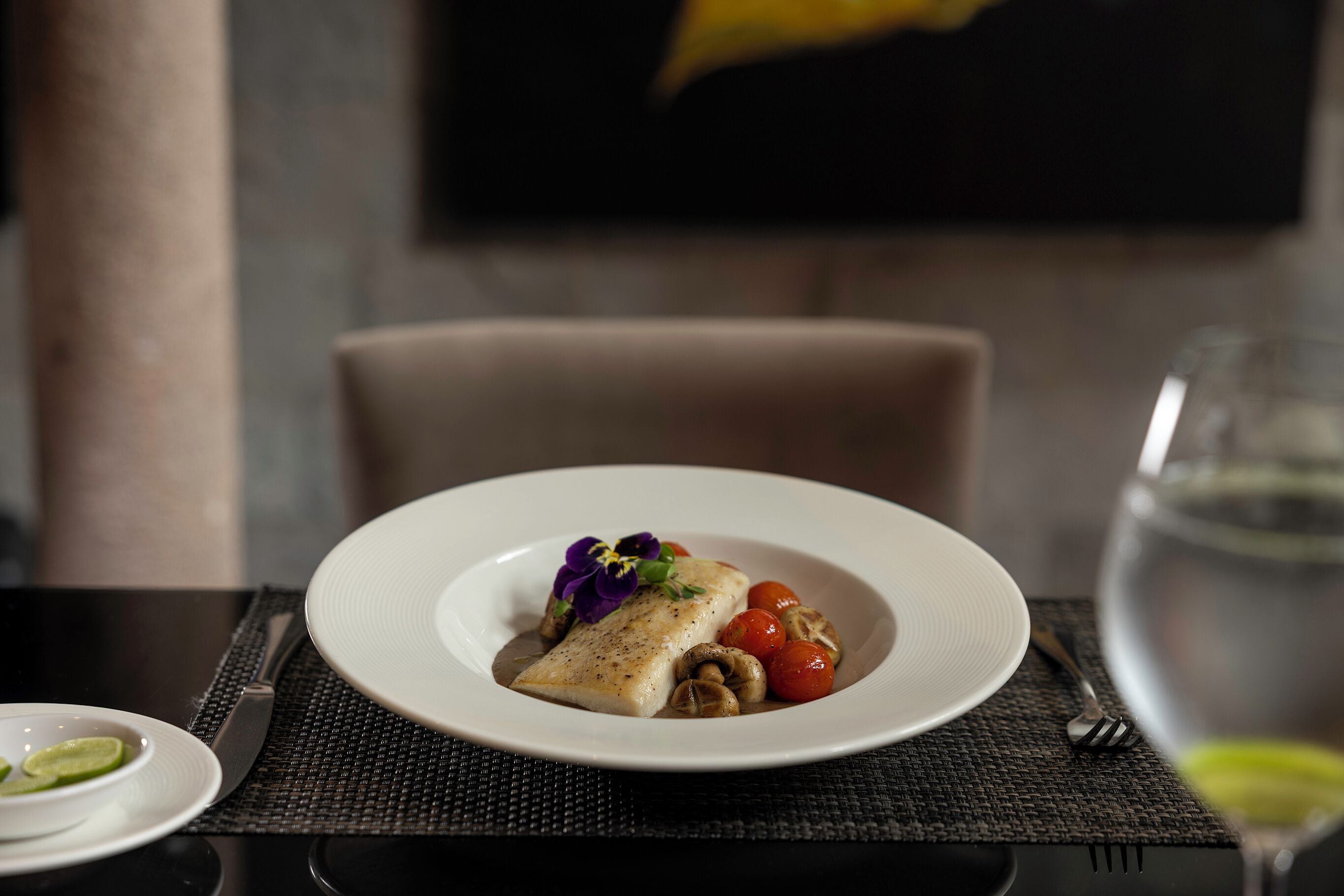 Um prato branco com filé de peixe, tomates cereja e uma flor roxa sobre uma mesa preta, com um garfo e uma faca ao lado.