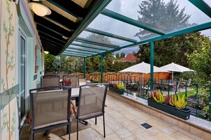 Uma área de varanda coberta com mobiliário externo, estrutura de vidro e vista para o jardim e árvores ao redor.
