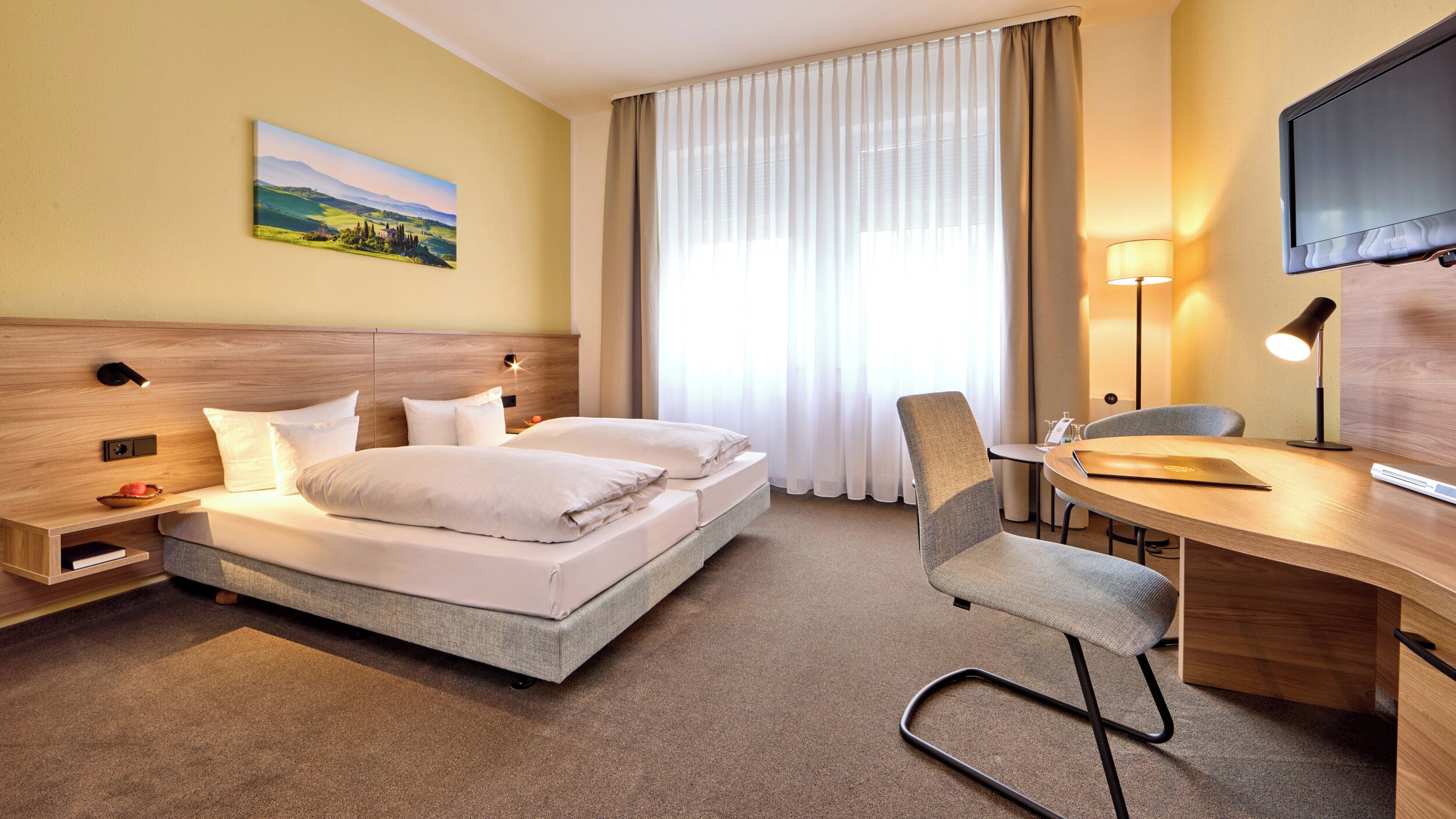 Quarto de hotel com cama, escrivaninha com cadeira, televisão e um quadro na parede.