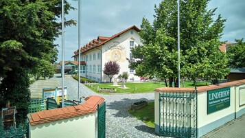 Um hotel com estilo arquitetônico europeu tradicional, com um caminho de pedras que leva à entrada, cercado por vegetação exuberante e um céu limpo.