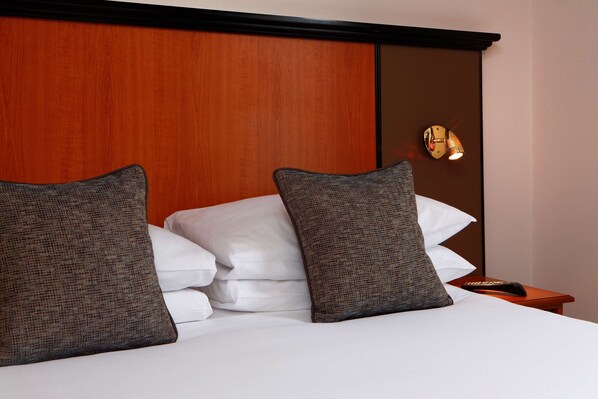 Cama bem arrumada com lençóis brancos e duas almofadas marrons escuras, cabeceira de madeira e uma pequena mesa de cabeceira com abajur.