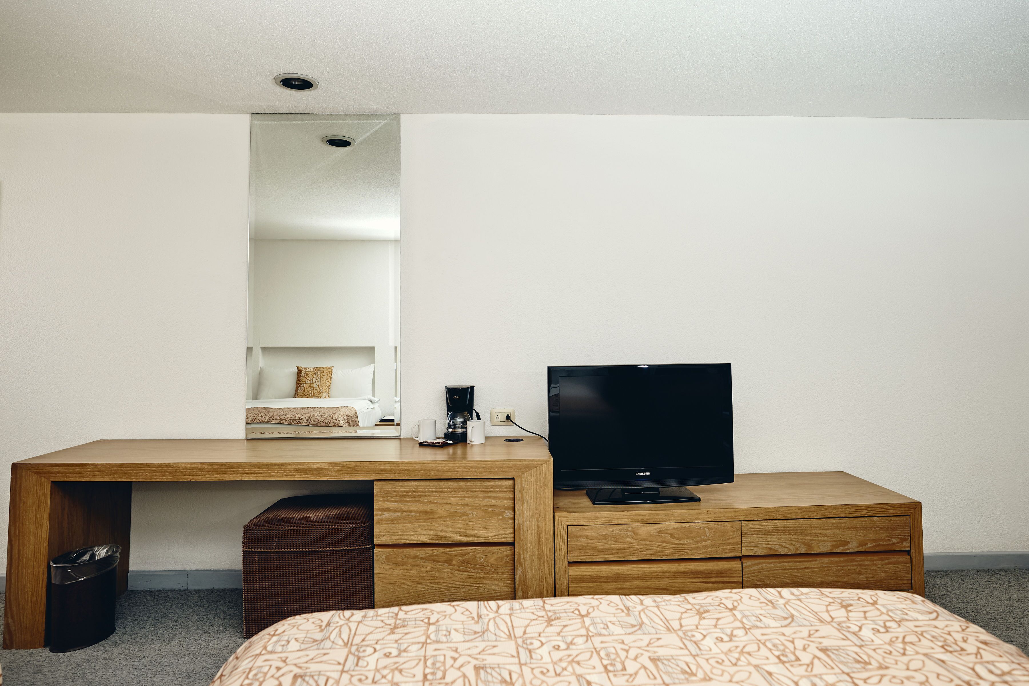 Quarto de hotel com escrivaninha de madeira, televisão, cama com roupa de cama estampada, cesto de vime pequeno e lixeira.