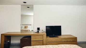 Quarto de hotel com escrivaninha de madeira, televisão, cama com roupa de cama estampada, cesto de vime pequeno e lixeira.