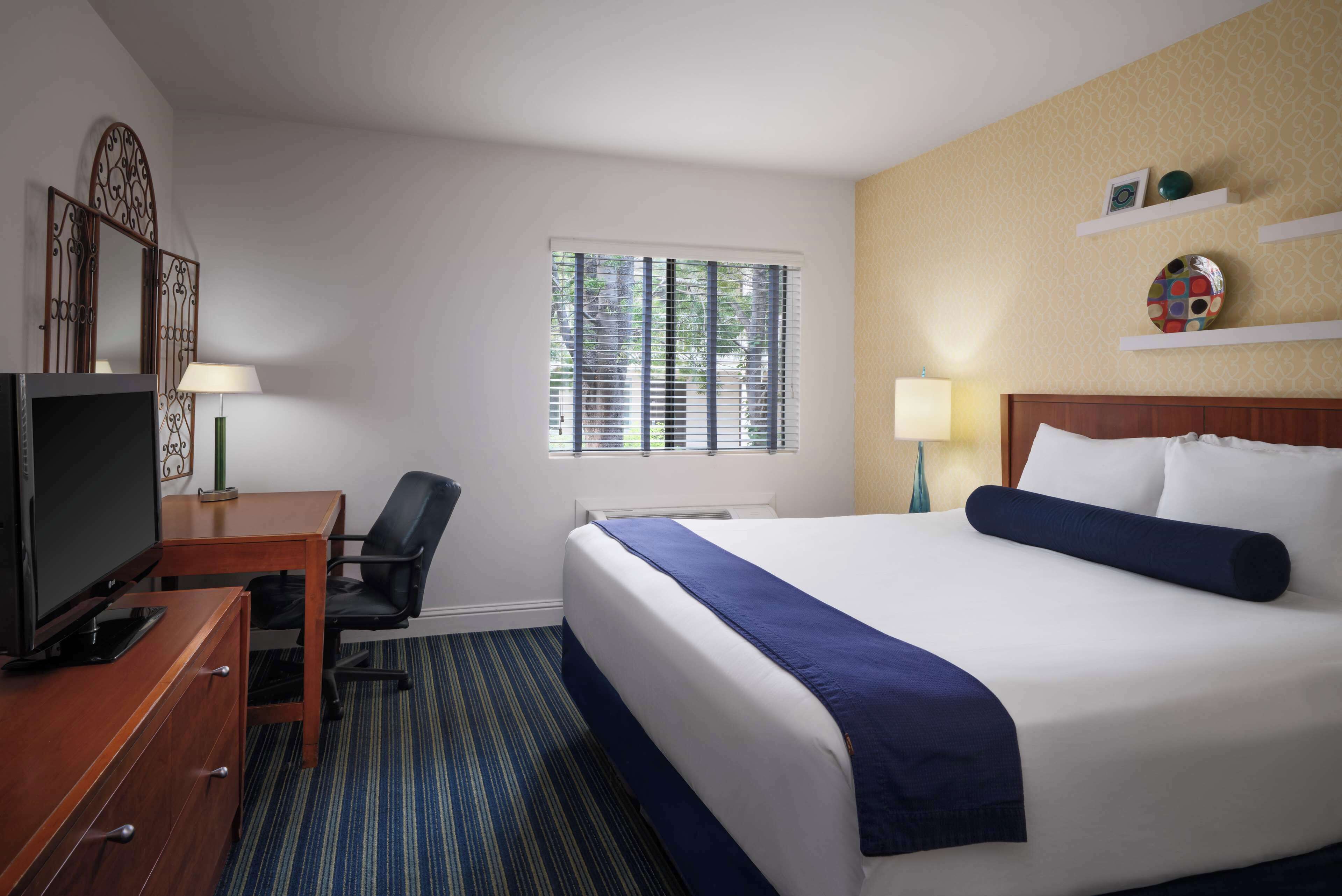 Quarto de hotel com cama, escrivaninha com computador, televisão, cadeira e janela com persianas.