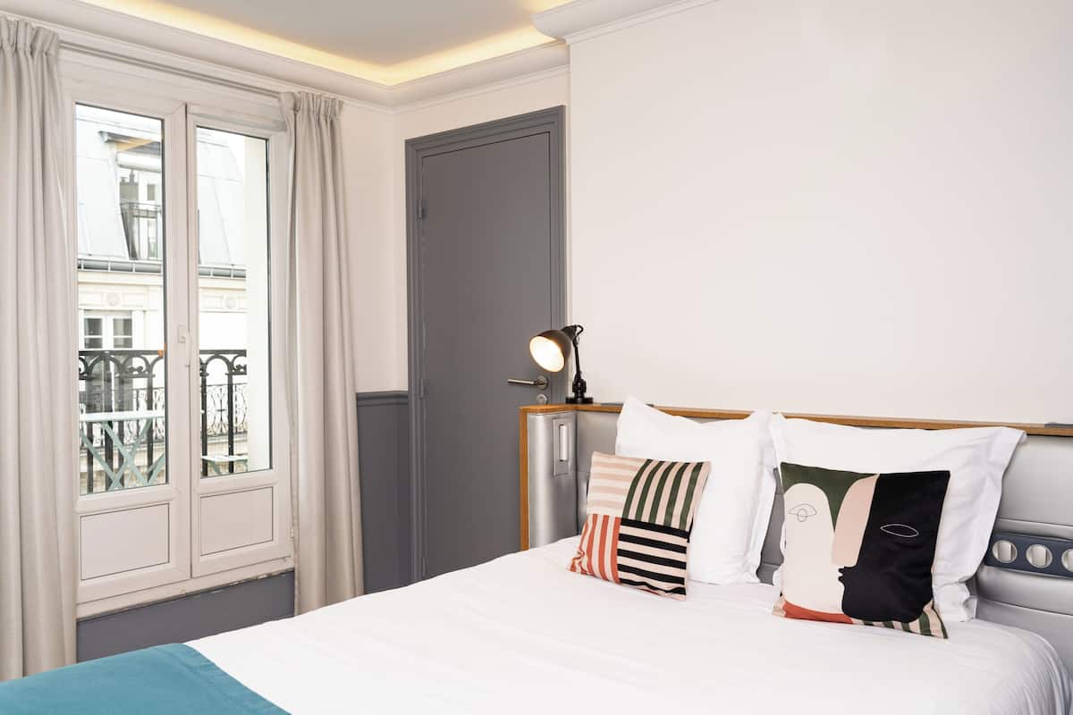 Quarto de hotel com cama, mesa de cabeceira, luminária e vista da cidade pela janela.