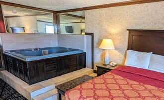 Quarto de hotel com uma banheira grande, uma cama com colcha estampada em vermelho e laranja, uma mesa de cabeceira com abajur e um espelho sobre a banheira.