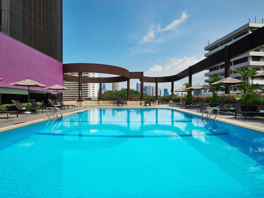 Uma piscina com espreguiçadeiras e guarda-sóis, cercada por edifícios modernos e um céu claro.