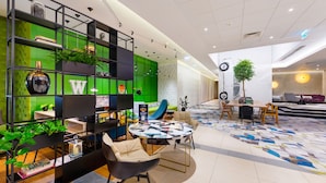 Um lobby de hotel moderno com uma parede verde de destaque, uma estante de metal preta, uma mesa redonda com cadeiras e um chão estampado.