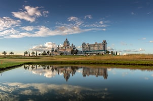 Um edifício grande e histórico refletido em águas calmas, rodeado por áreas gramadas e um céu claro.