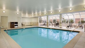 Uma piscina interior com água cristalina, rodeada por espreguiçadeiras e mesas, com amplas janelas que oferecem vista para o exterior.